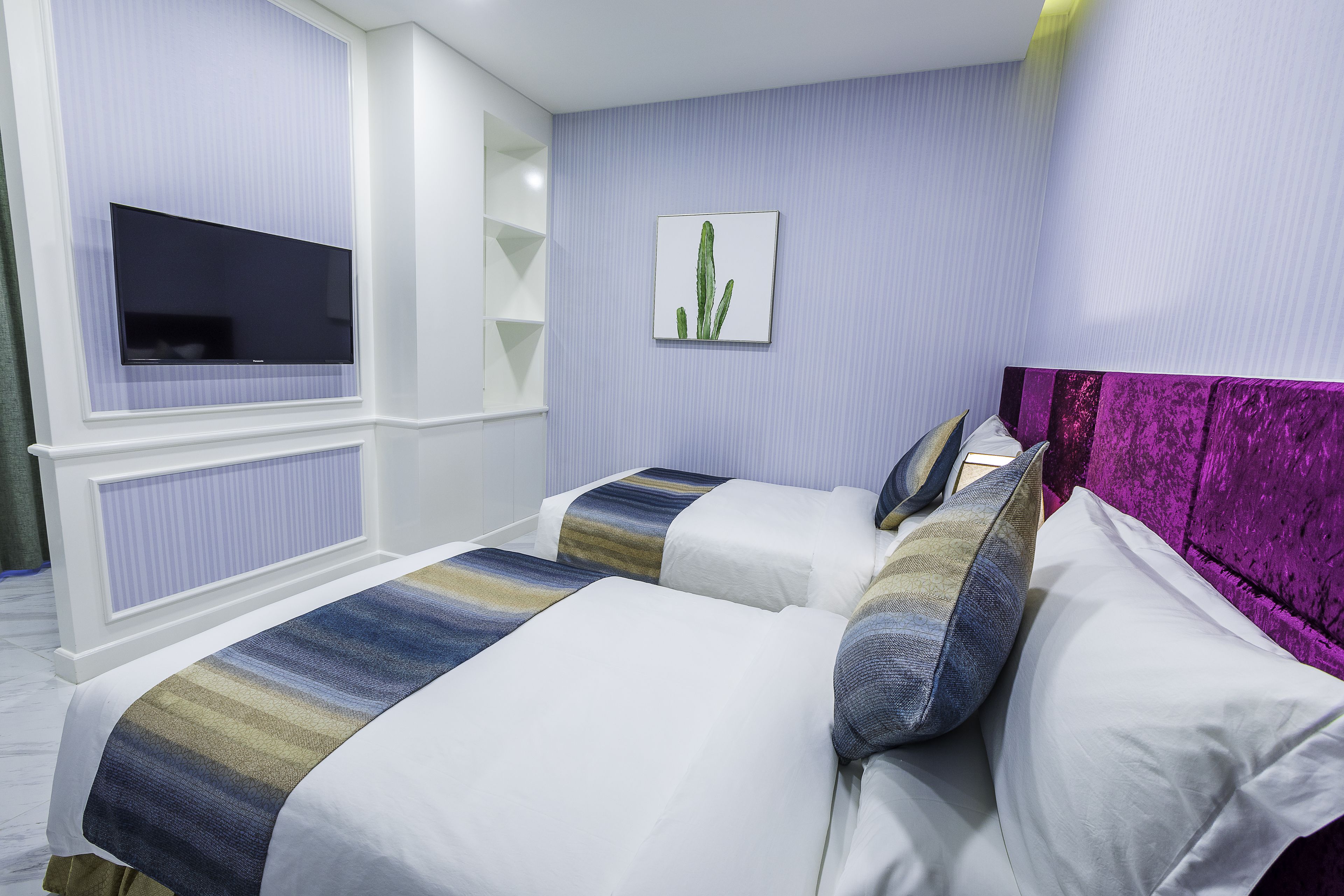 Quarto de hotel com duas camas, televisão, obra de arte de cacto e cabeceira roxa.