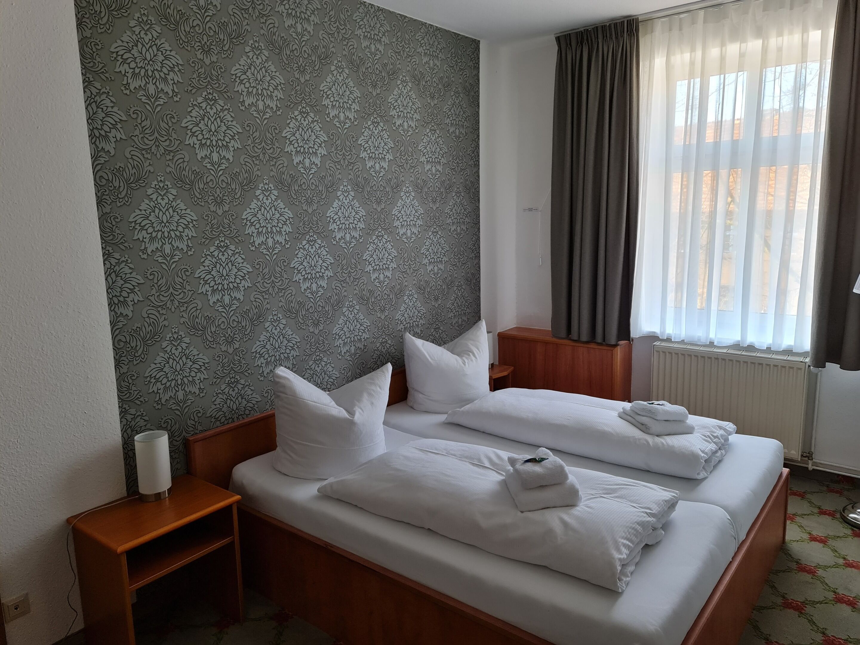 Quarto de hotel com duas camas, cabeceira de madeira, mesa de cabeceira, luminária e janela com cortinas.