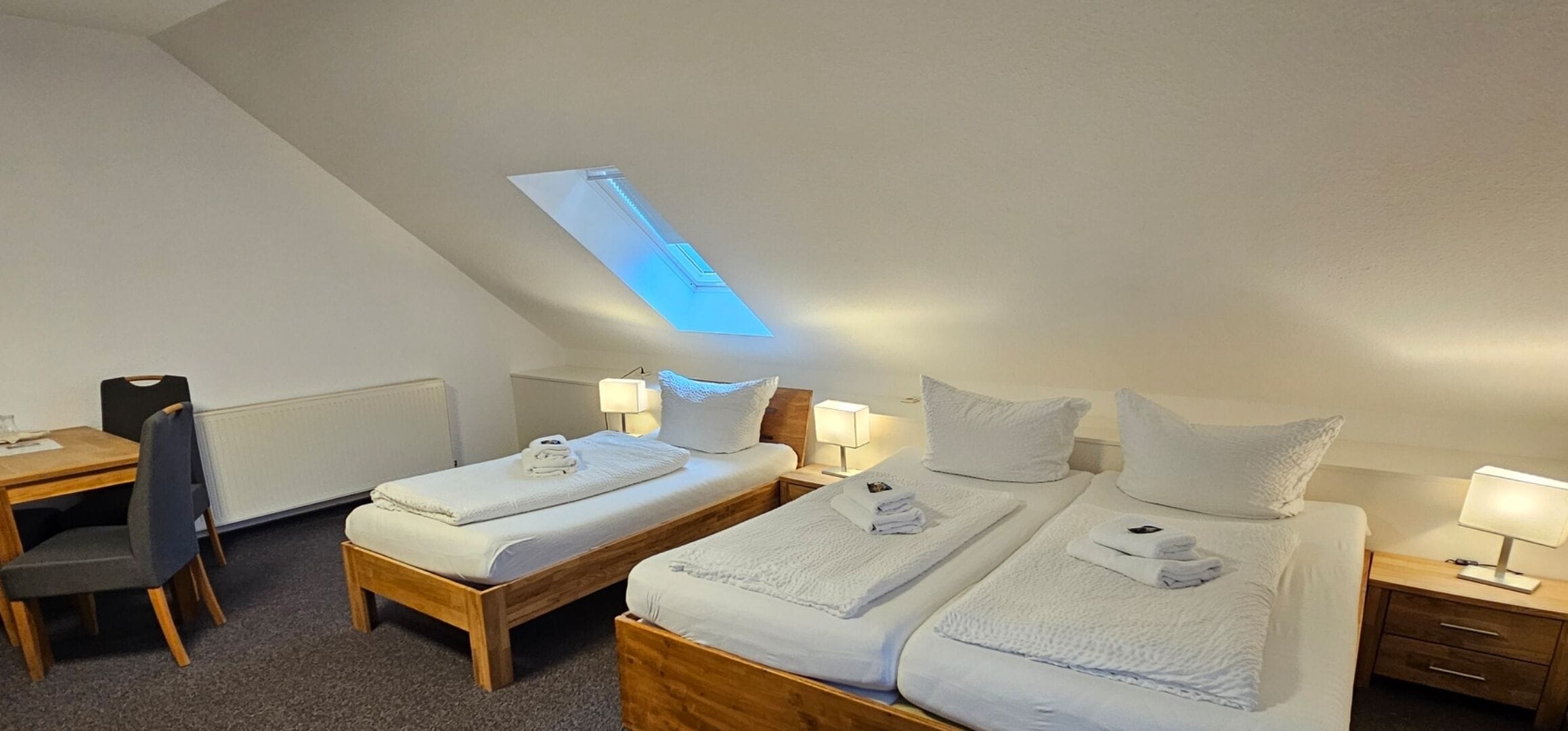 Quarto de hotel com duas camas de solteiro, uma escrivaninha, uma cadeira e um claraboia.