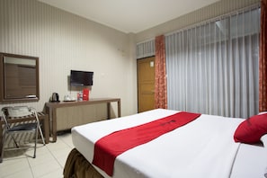 Quarto de hotel com cama, escrivaninha com televisão, cadeira e janela com cortinas.