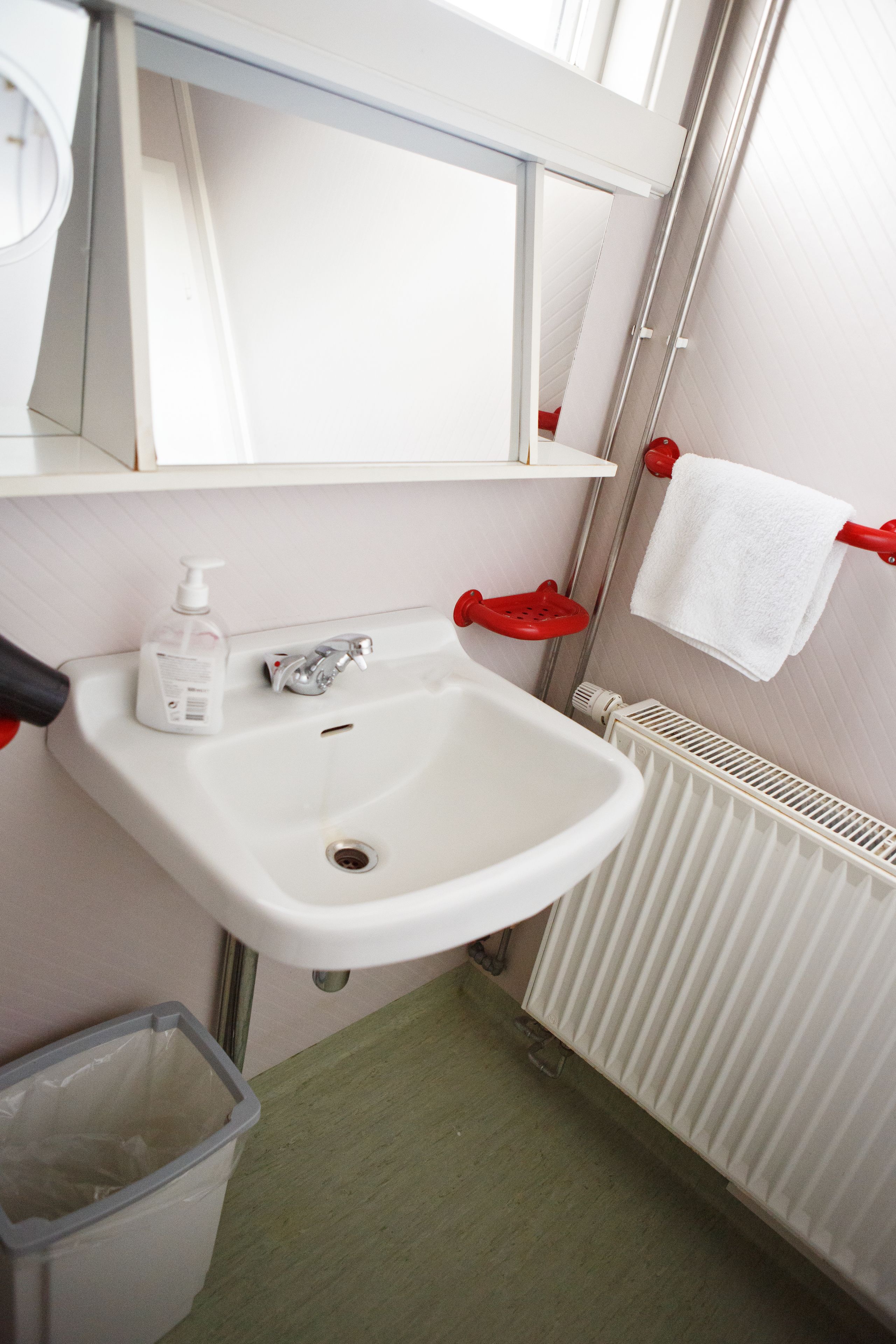 Casa de banho com uma pia branca, um espelho, um porta-toalhas vermelho, um radiador branco e um cesto de lixo.
