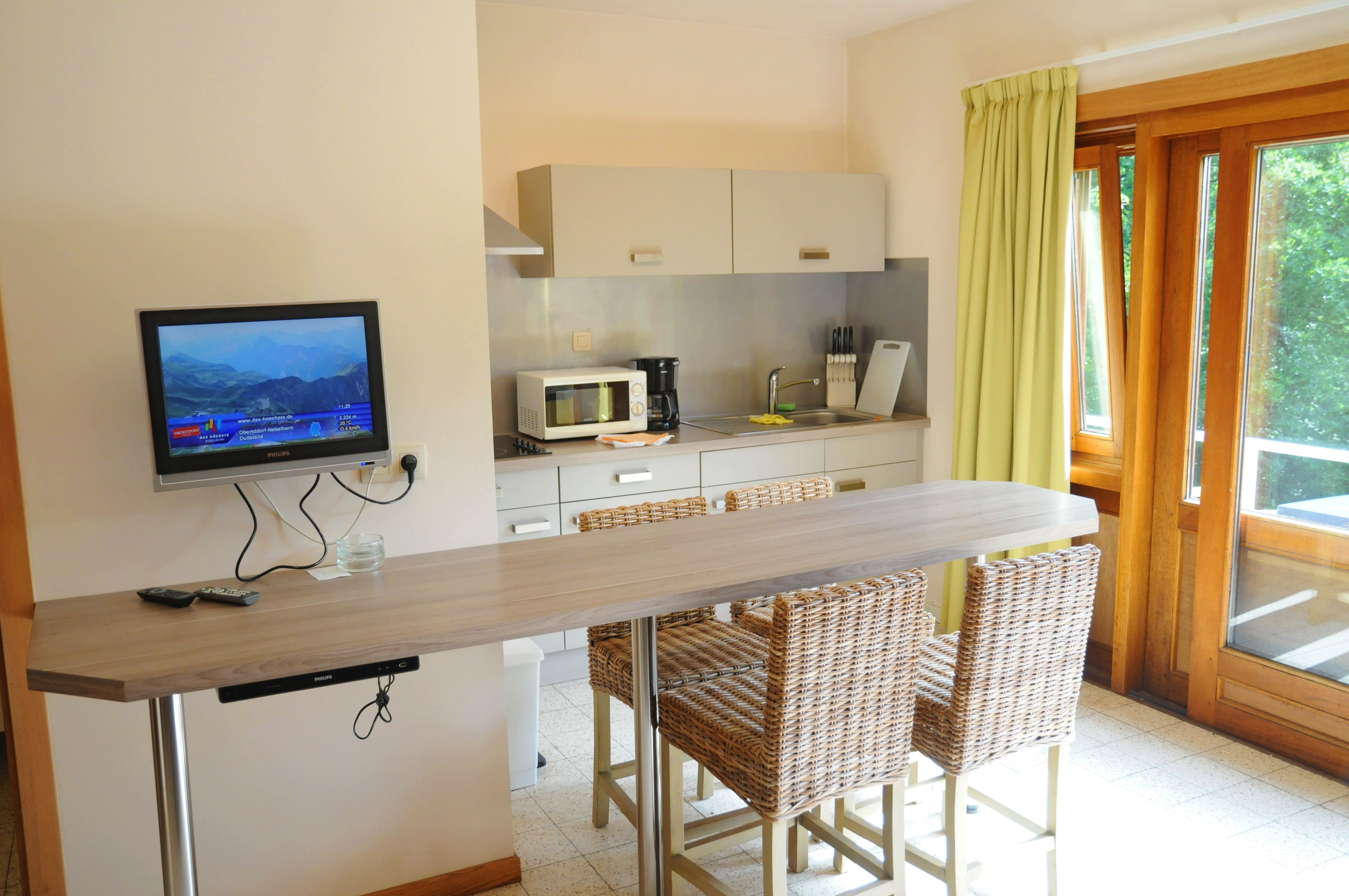 Uma cozinha moderna com televisão de tela plana, micro-ondas e cafeteira. Há uma área de refeições com cadeiras de vime e mesa de madeira.