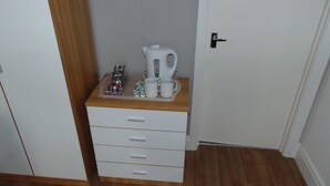 Um quarto pequeno com um armário de madeira, uma chaleira branca, duas chávenas brancas e um tabuleiro com alguns objetos em cima.
