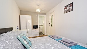 Quarto com cama, guarda-roupa, escrivaninha, televisão e banheiro visível por uma porta aberta.