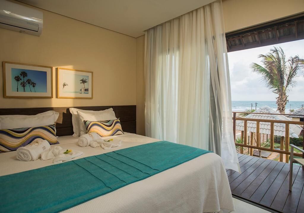 Quarto de hotel com cama, toalhas, vista para a praia e duas fotos emolduradas na parede.