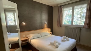 Quarto de hotel com cama, mesas de cabeceira, janela com cortinas, radiador e luminária no teto.