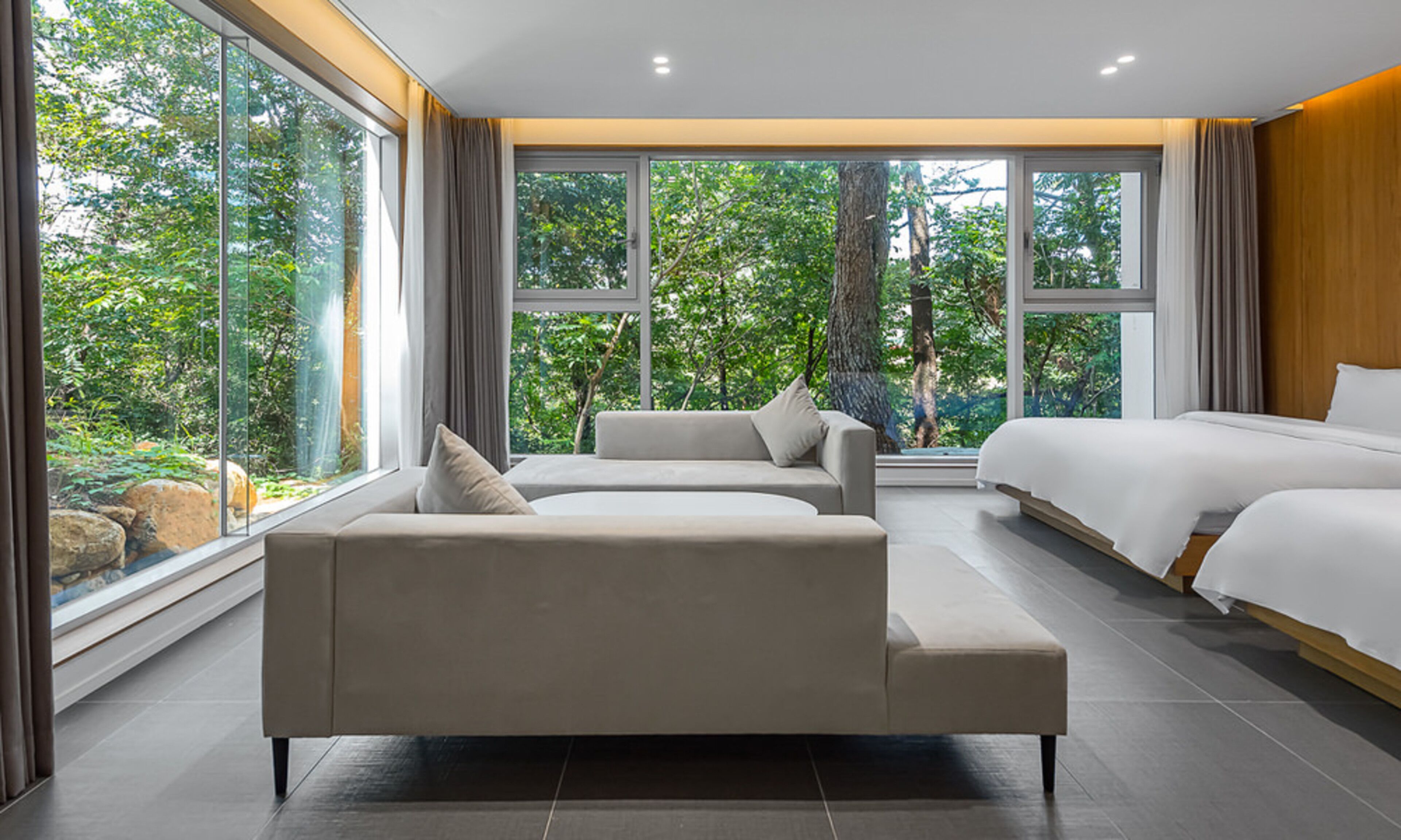 Quarto de hotel moderno com uma cama grande, um sofá e vista para uma floresta através de amplas janelas.