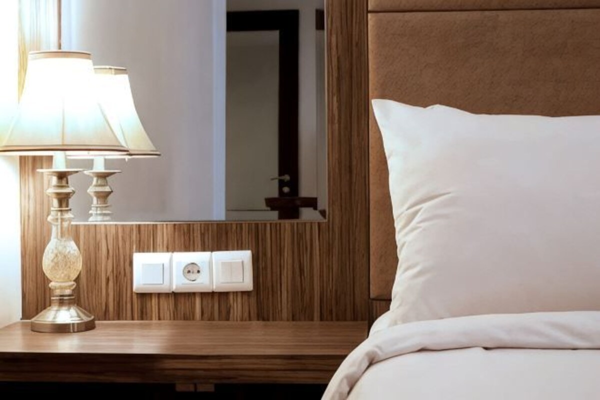 Quarto de hotel com cabeceira de madeira, luminária de mesa, interruptor junto à cama e espelho.