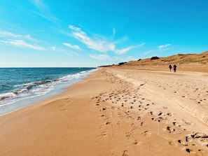 Uma extensa praia de areia com pegadas que se estendem em direção à água, sob um céu azul claro.