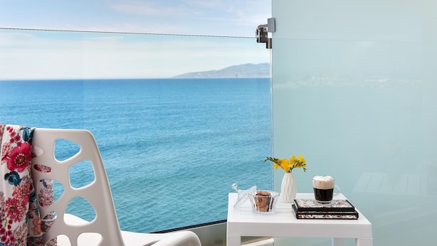Quarto com vista para o mar, uma cadeira branca com capa florida, uma mesa pequena com um vaso de flores amarelas, uma garrafa de vidro e uma caneca.
