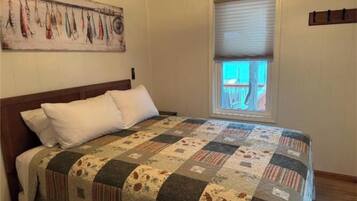 Quarto com cabeceira de madeira, colcha acolchoada, janela com vista para uma varanda e uma obra de arte na parede com iscas de pesca.