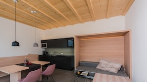 Quarto compacto com teto de madeira, cozinha compacta, cama com roupa de cama estampada e área de refeições com mesa e cadeiras.