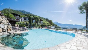 Piscina com ladrilhos de pedra, rodeada por espreguiçadeiras e guarda-sóis, com montanhas ao fundo.