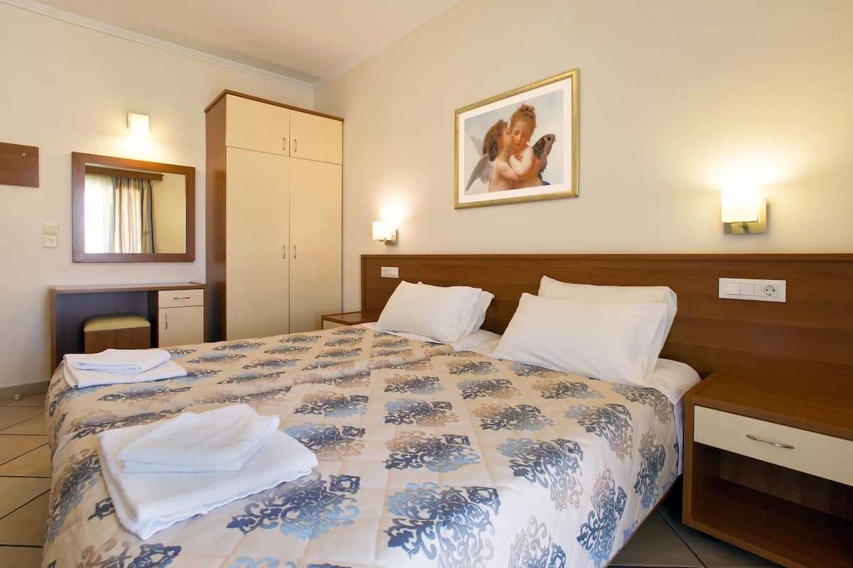Quarto de hotel com uma cama grande, cabeceira de madeira, mesa de cabeceira, escrivaninha, guarda-roupa e um quadro emoldurado na parede.