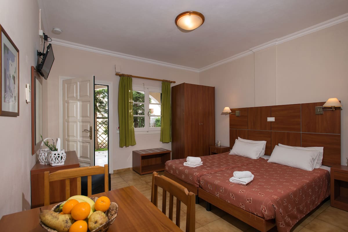 Quarto de hotel com uma cama, uma mesa com uma cesta de frutas, uma cadeira de madeira, uma mesinha de cabeceira e uma janela com cortinas.