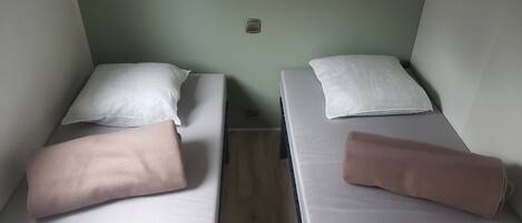 Duas camas de solteiro com roupa de cama branca e rosa, uma luminária pequena na parede e piso de madeira.