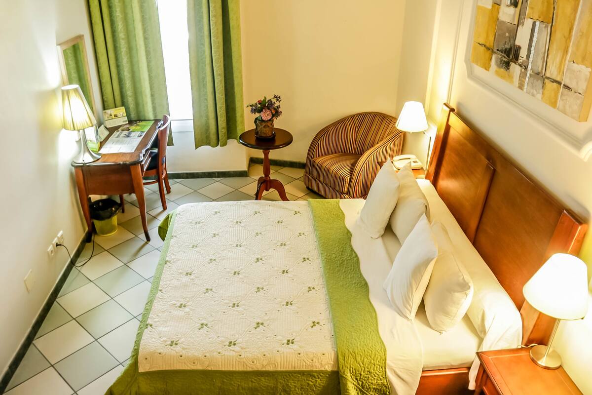 Quarto de hotel com cama, escrivaninha, cadeira, mesa pequena com um vaso e um quadro na parede.
