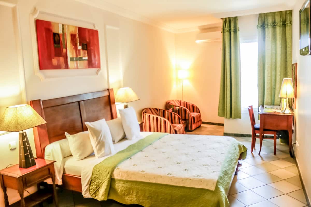 Quarto de hotel com cama, duas luminárias de cabeceira, uma cadeira, uma escrivaninha e janela com cortinas.