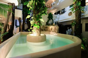 Uma grande fonte interior com uma árvore central, rodeada por vegetação exuberante e paredes de pedra.