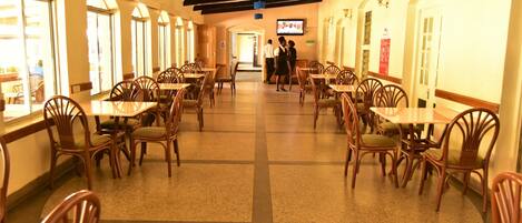 Restaurante com cadeiras e mesas de madeira, uma tela de televisão e chão de azulejos.