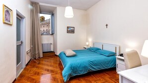 Quarto com cama, escrivaninha, cadeira, janela com vista para um edifício e luminária pendente.
