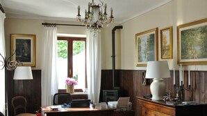 Quarto tradicional com escrivaninha de madeira, uma cadeira, cômoda e lustre. Há quadros nas paredes e uma janela com cortinas.
