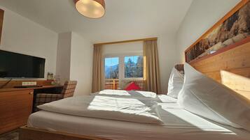Quarto de hotel com cama, escrivaninha com televisão e janela com vista para árvores e montanhas.