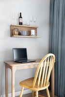 Uma mesa de madeira com um laptop, uma cadeira de madeira, uma estante com copos de vinho, uma garrafa de vinho e uma xícara de café.