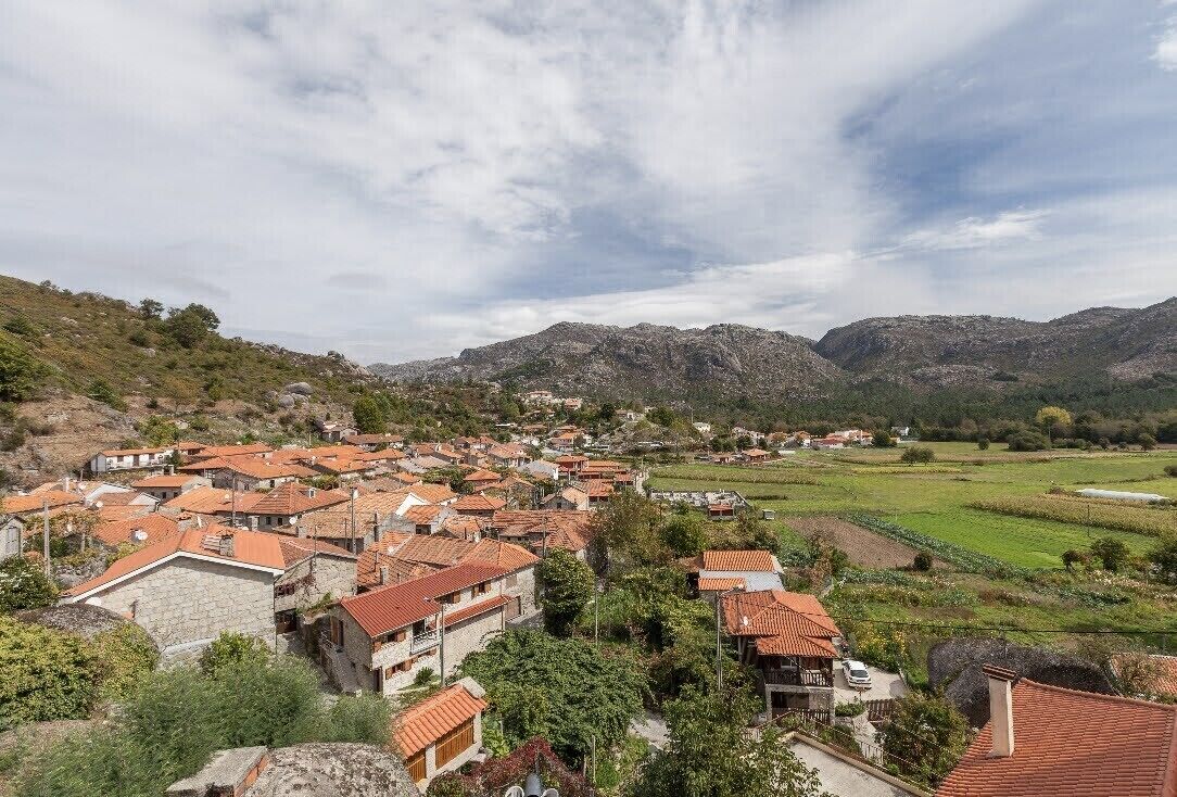 Uma aldeia com edifícios de telhados em terracota, rodeada por campos verdes e montanhas.