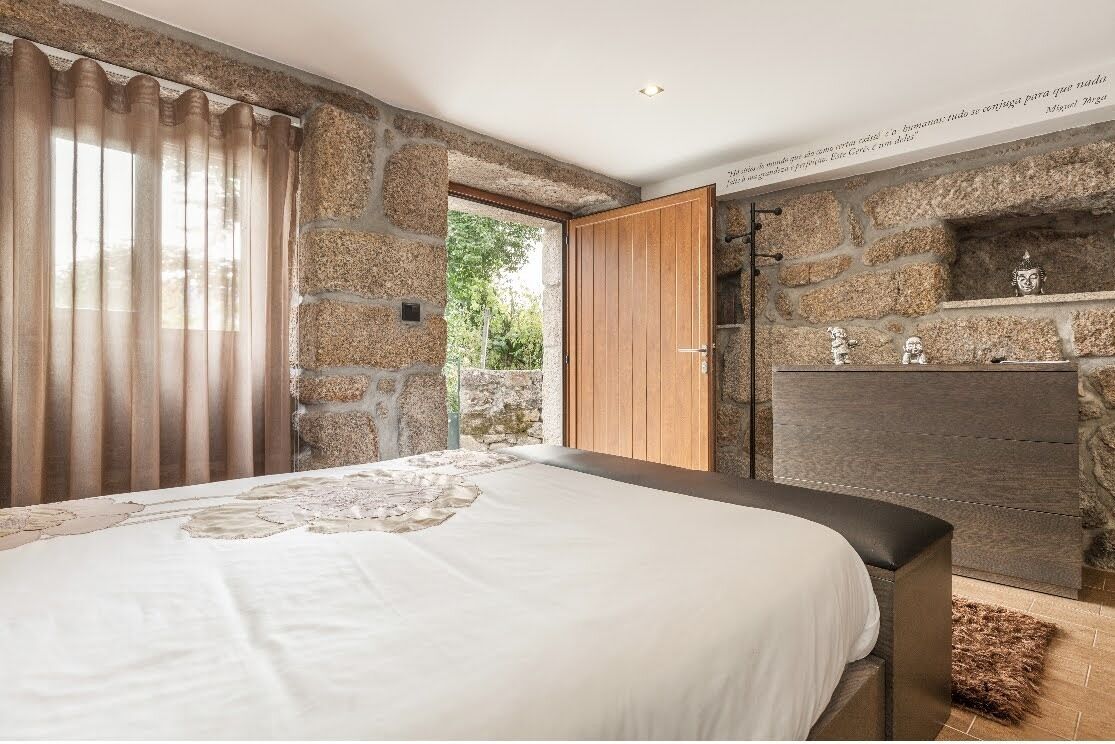 Quarto com parede de pedra, cama com colcha branca e porta de madeira.