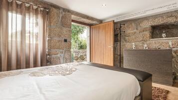 Quarto com parede de pedra, cama com colcha branca e porta de madeira.