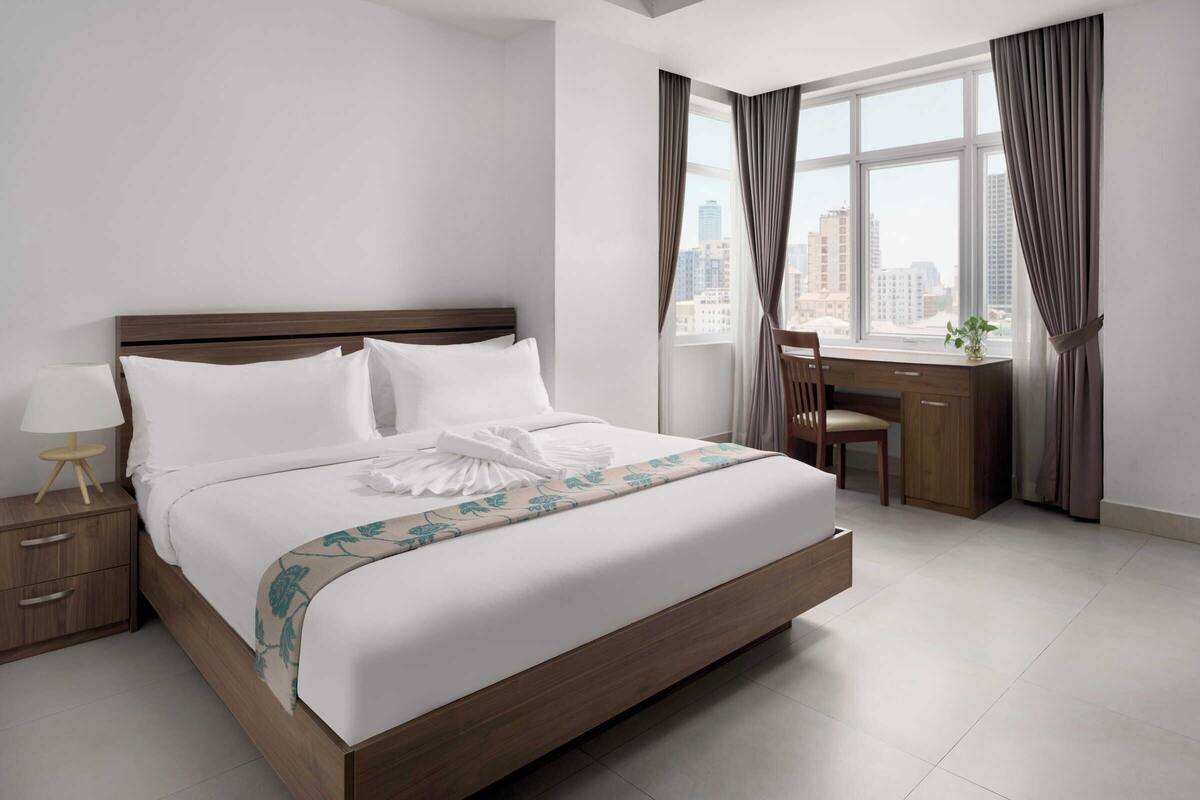 Cama bem arrumada com lençóis brancos, cabeceira de madeira e manta com padrão floral. Há uma escrivaninha com cadeira, uma janela com cortinas e uma mesa de cabeceira com luminária e gavetas.