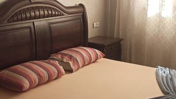 Quarto com cabeceira de madeira, cama com almofadas listradas, manta cinza e janela com cortinas leves.