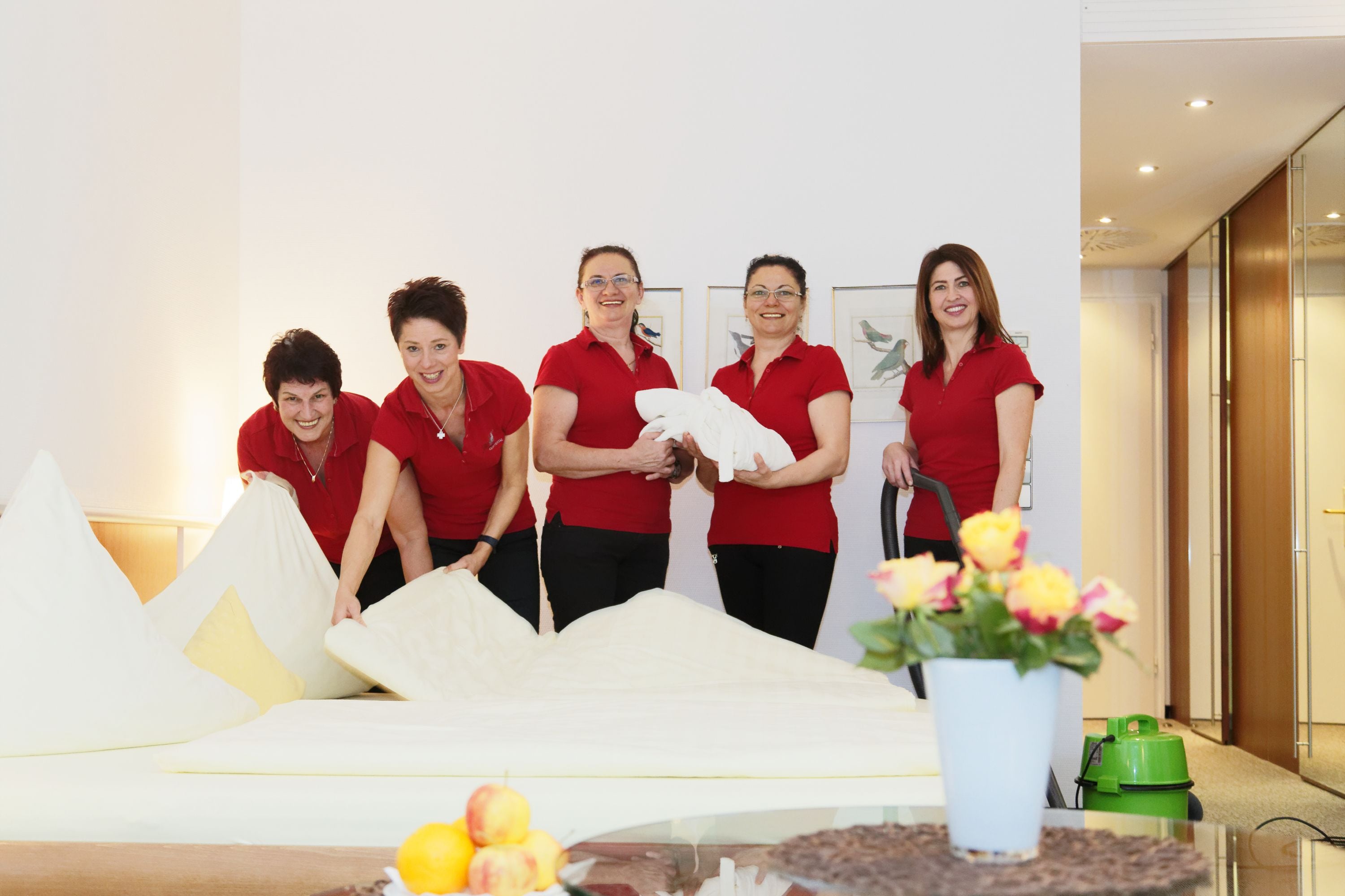 Um grupo de pessoas de camisas vermelhas arrumando uma cama em quarto de hotel, com um vaso de flores na mesa.