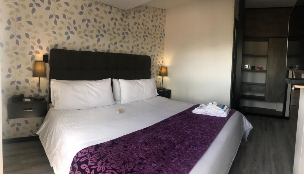 Quarto de hotel com uma cama, duas mesas de cabeceira, um criado-mudo, um cobertor roxo e uma parede com padrão de folhas.