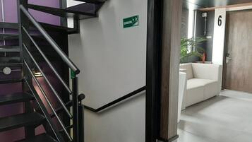 Um interior moderno com escadas, uma placa indicando saída de emergência, uma área de recepção com sofá e uma porta marcada com o número 6.
