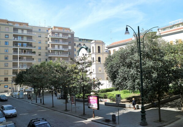 Uma rua com edifícios residenciais de vários andares, uma igreja com uma torre de sino e árvores ao longo da calçada.