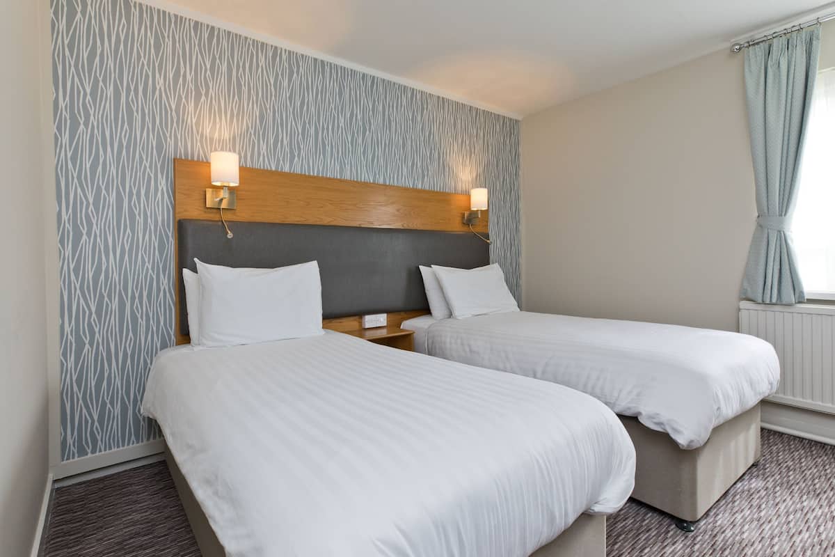 Quarto de hotel com duas camas de solteiro, cabeceira de madeira, papel de parede estampado, mesa de cabeceira e uma janela com cortinas.