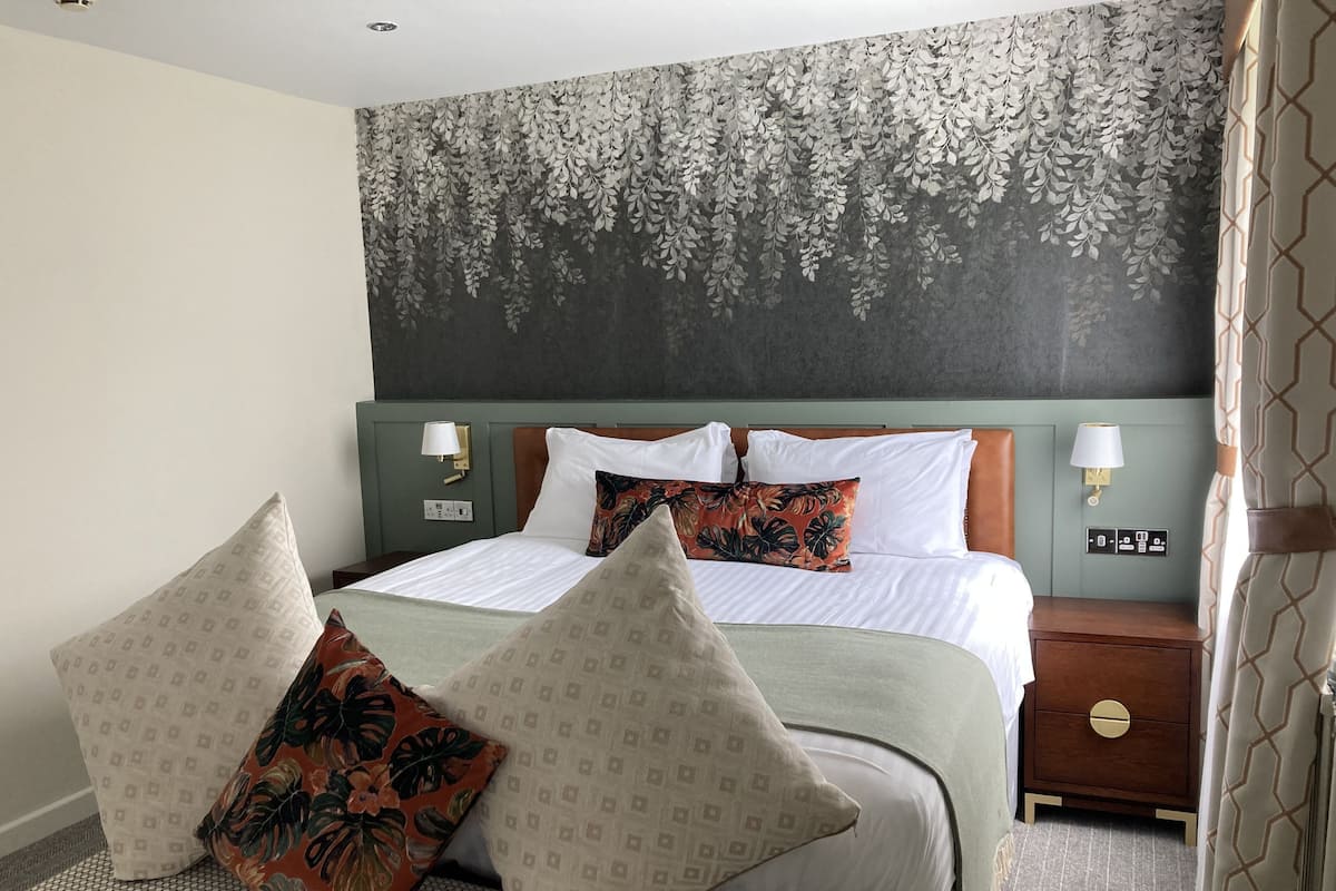 Quarto de hotel com uma cama, duas mesas de cabeceira, uma luminária na parede e cortina estampada.
