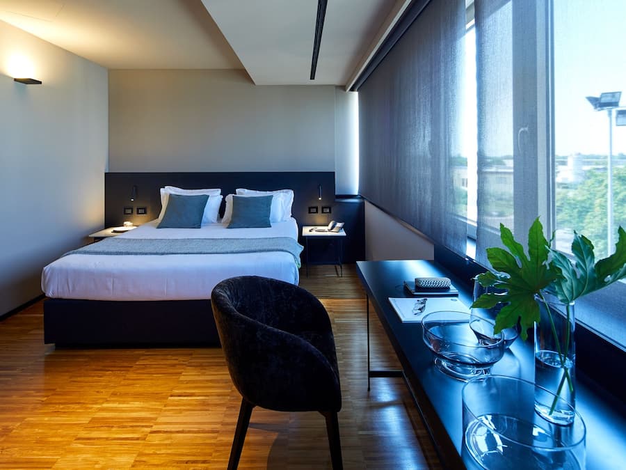 Quarto de hotel moderno com uma cama grande, uma escrivaninha com cadeira, uma janela com vista e uma planta no parapeito.