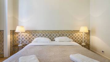 Uma cama bem arrumada com lençóis brancos e duas luminárias de cabeceira, um painel de cama com padrão floral e uma pequena mesa de cabeceira com um abajur.