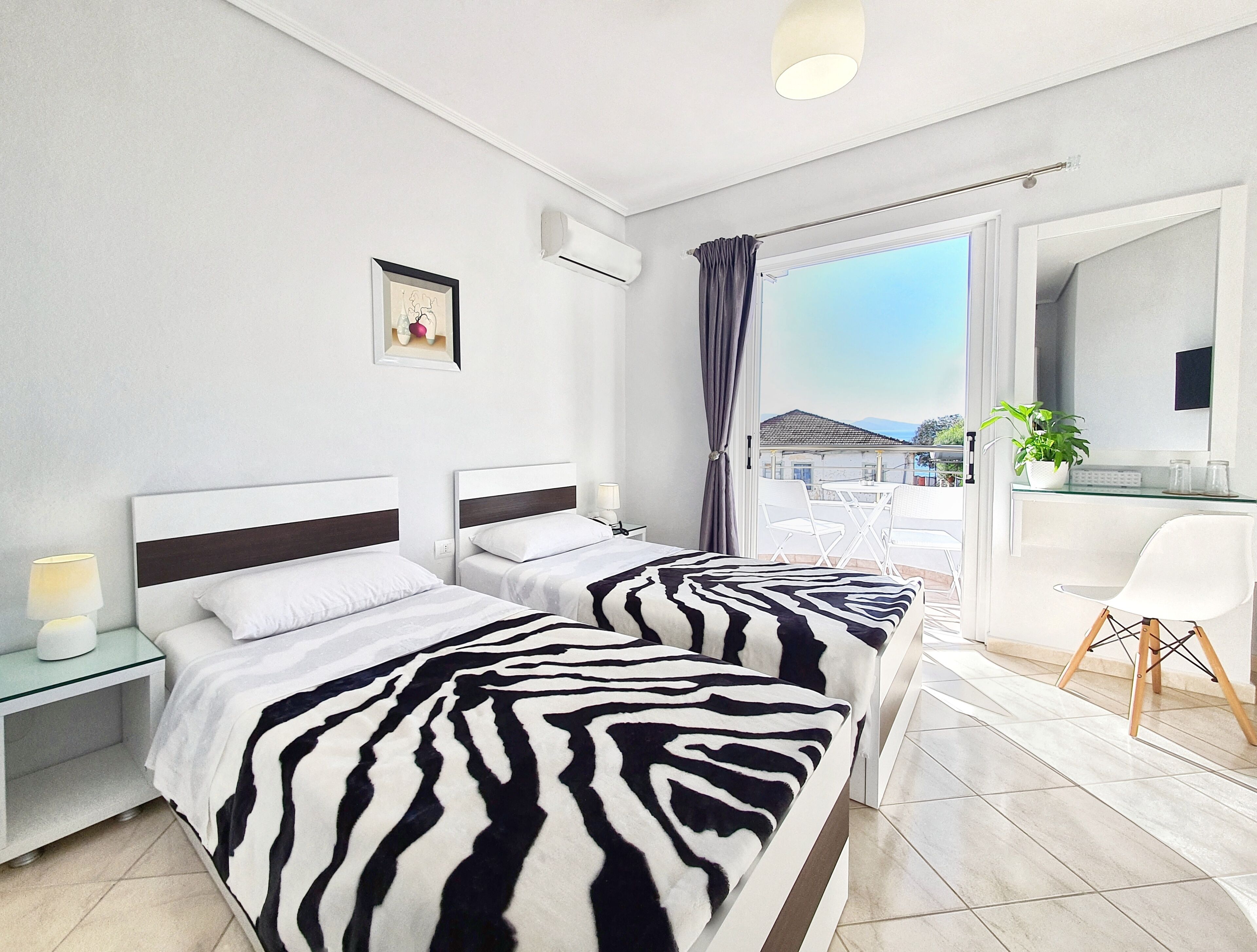Quarto de hotel com duas camas, varanda com cadeiras, um vaso de planta e vista para a cidade.