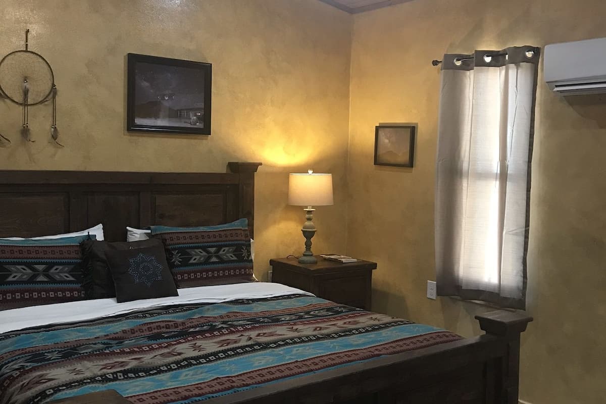 Quarto com cama de madeira, mesa de cabeceira com luminária, ar condicionado fixado na parede e janela com cortinas.