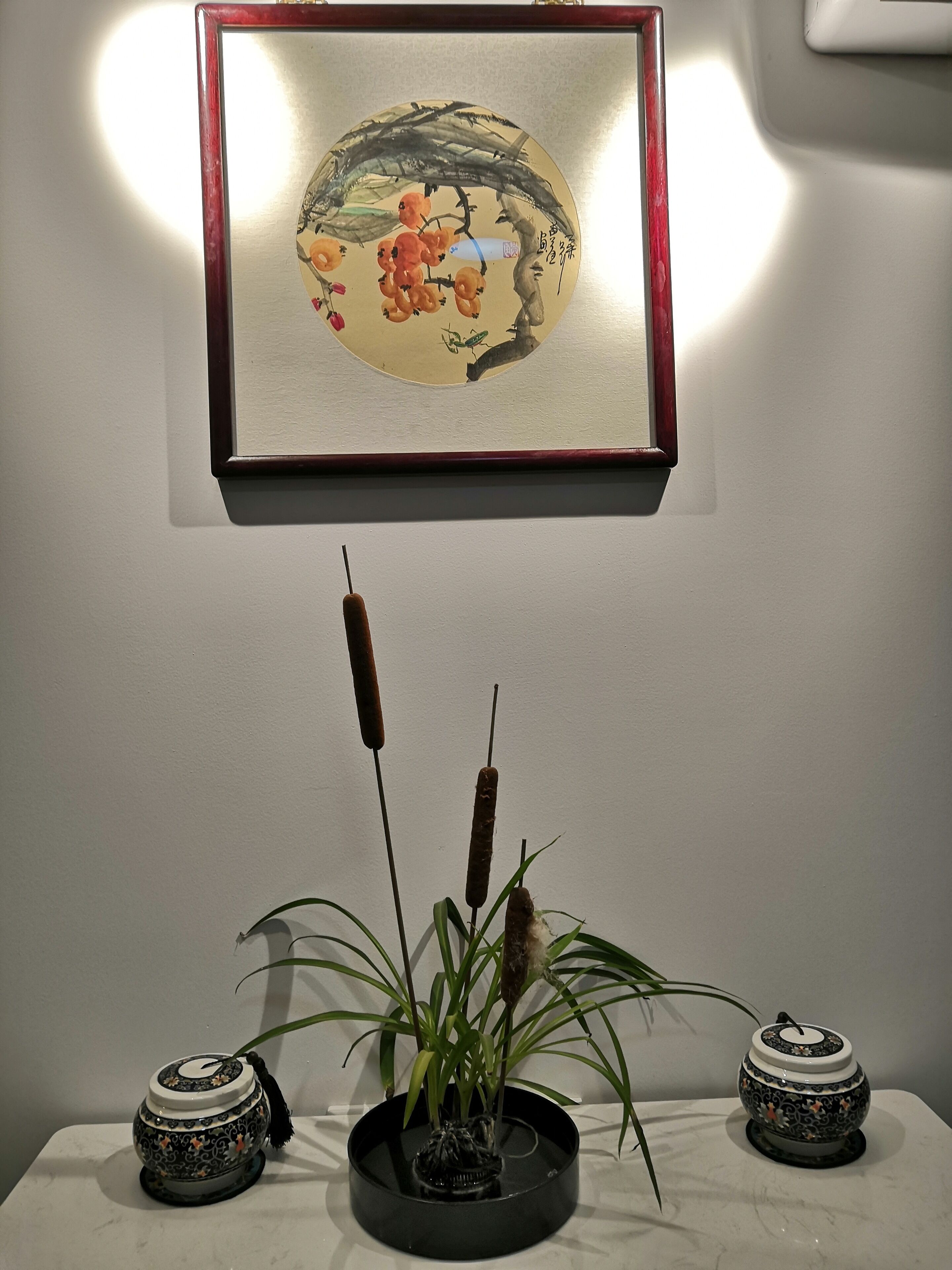 Uma obra de arte emoldurada retratando uma árvore com frutos e insetos, uma planta em vaso com gramíneas altas e duas tigelas decorativas sobre uma mesa.