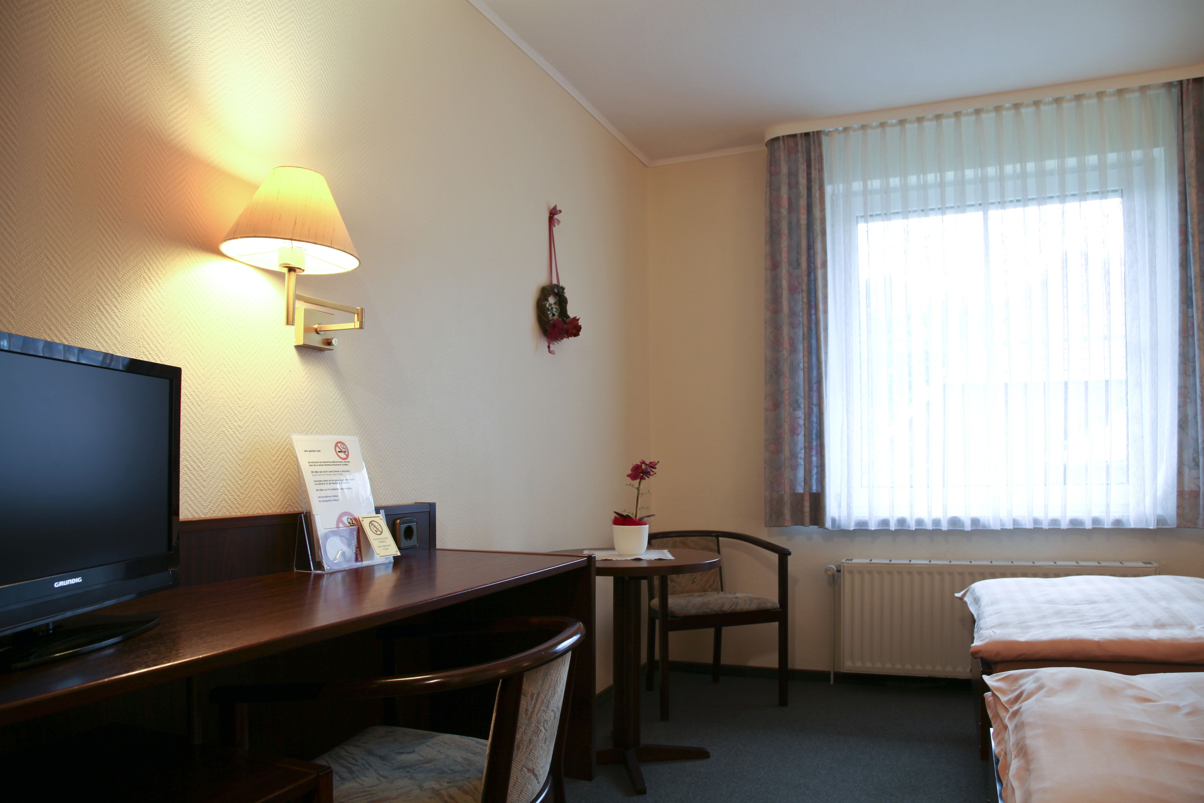 Quarto de hotel com cama, escrivaninha com televisão, cadeira, mesinha com uma flor e janela com cortinas.