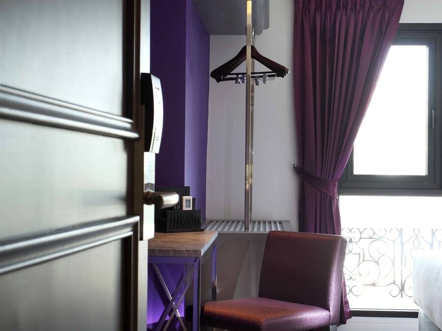 Quarto de hotel com piso de madeira, parede roxa, cama com cabeceira branca, escrivaninha com cadeira e janela com cortinas roxas.