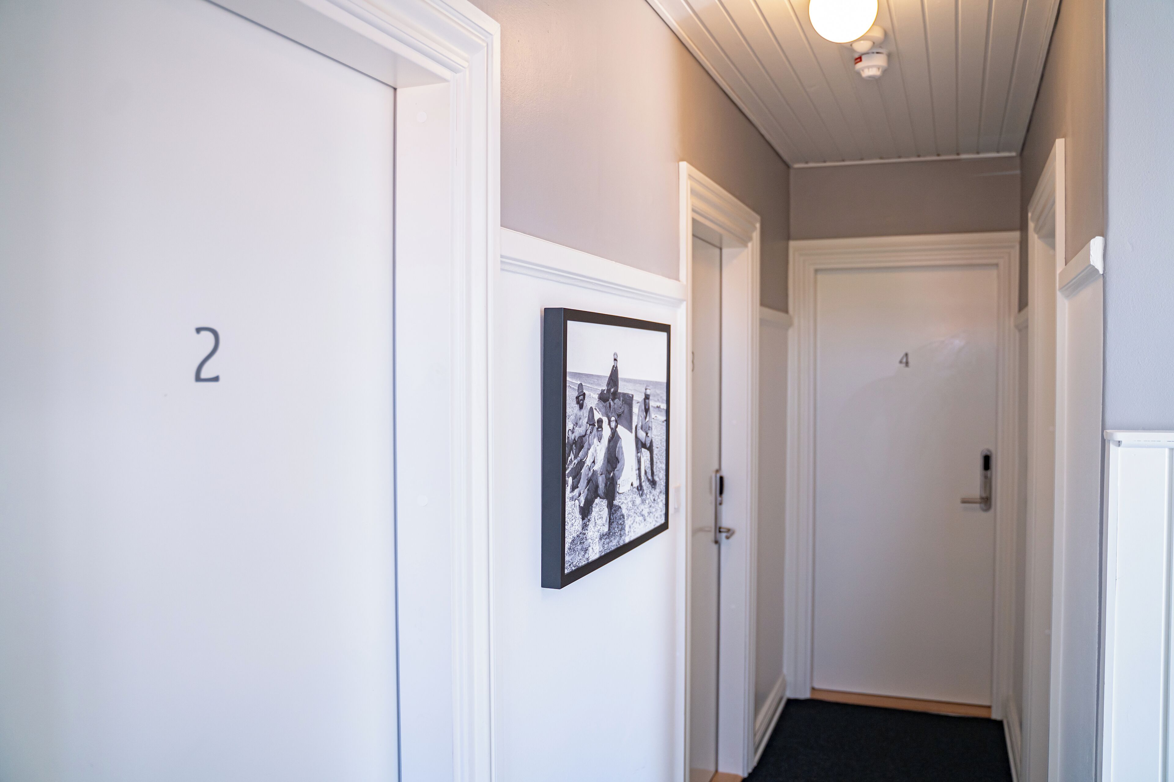 Um corredor com portas numeradas 2 e 4, uma fotografia em preto e branco emoldurada na parede e um luminário no teto.