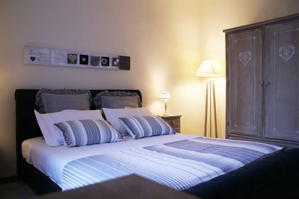 Cama bem arrumada com roupa de cama listrada, luminária de cabeceira, colagem de fotos na parede e guarda-roupa com detalhes em formato de coração.