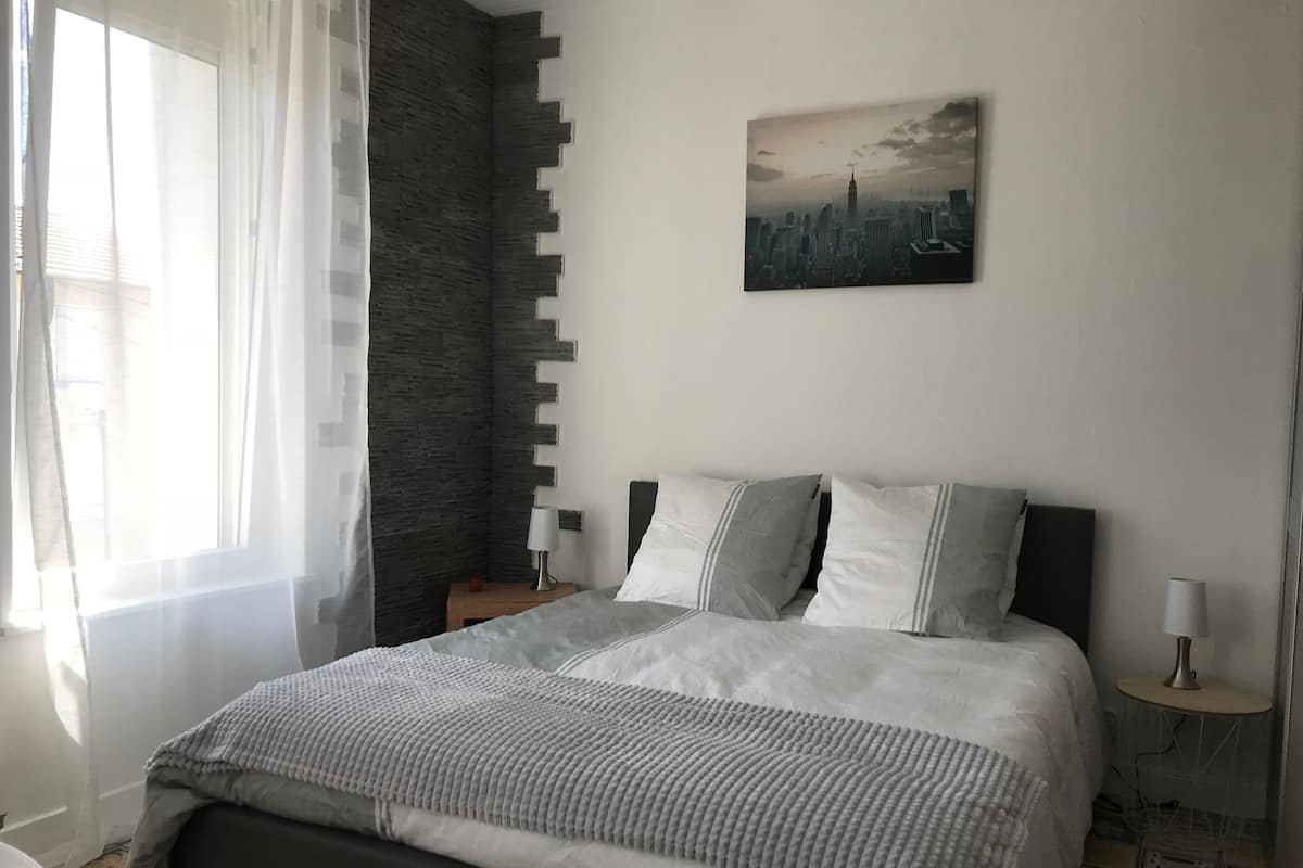 Quarto moderno com cama, pintura de paisagem urbana e janela com cortinas leves.