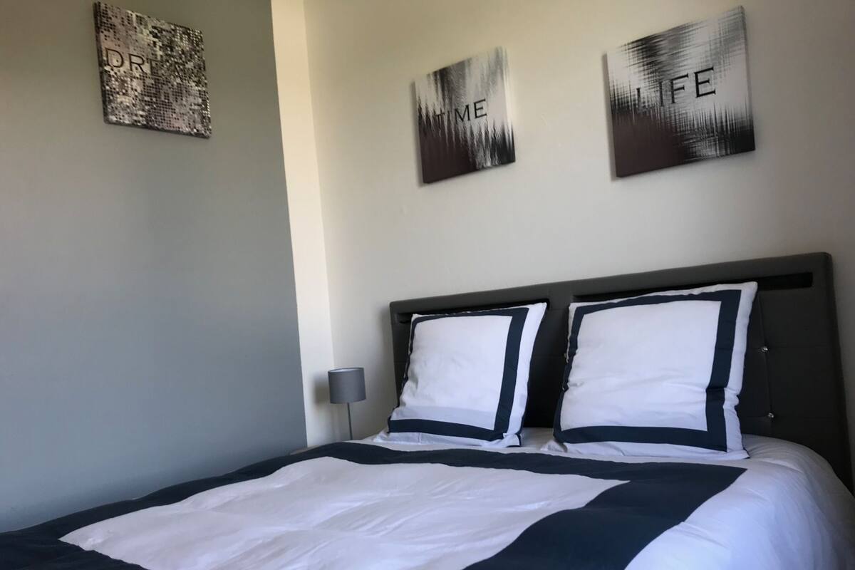 Cama bem arrumada com roupa de cama branca e escura, dois travesseiros e cabeceira. Três obras de arte penduradas na parede com texto.